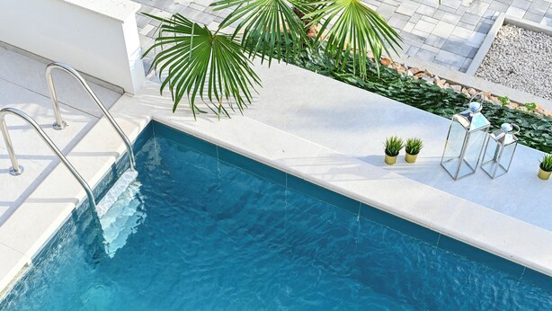 Piscina com escada de metal, rodeada por vasos de plantas e um deck revestido de azulejos.