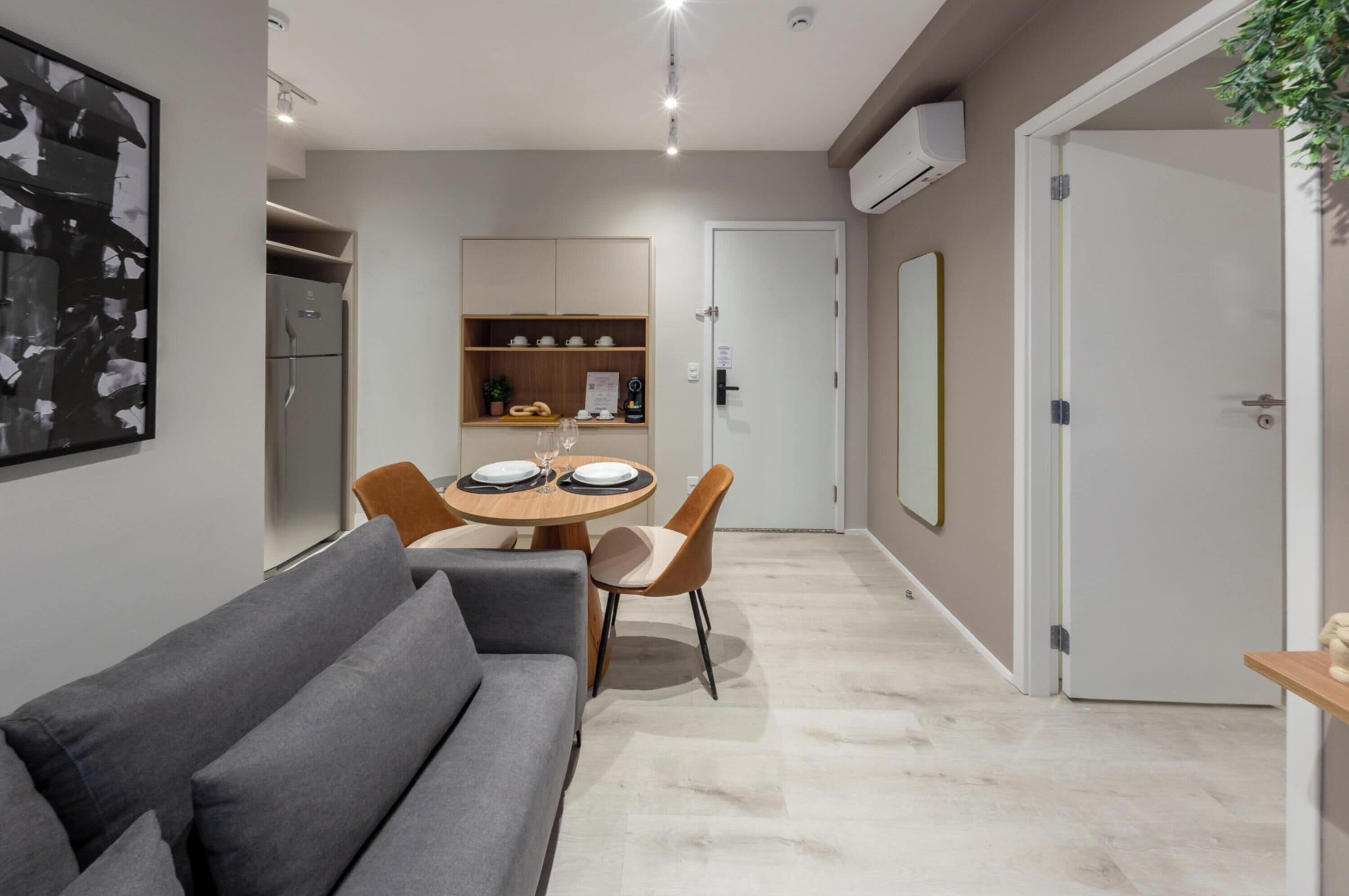 Uma sala de estar moderna com um sofá cinzento, uma mesa de jantar com cadeiras e uma área de cozinha com frigorífico e estantes.