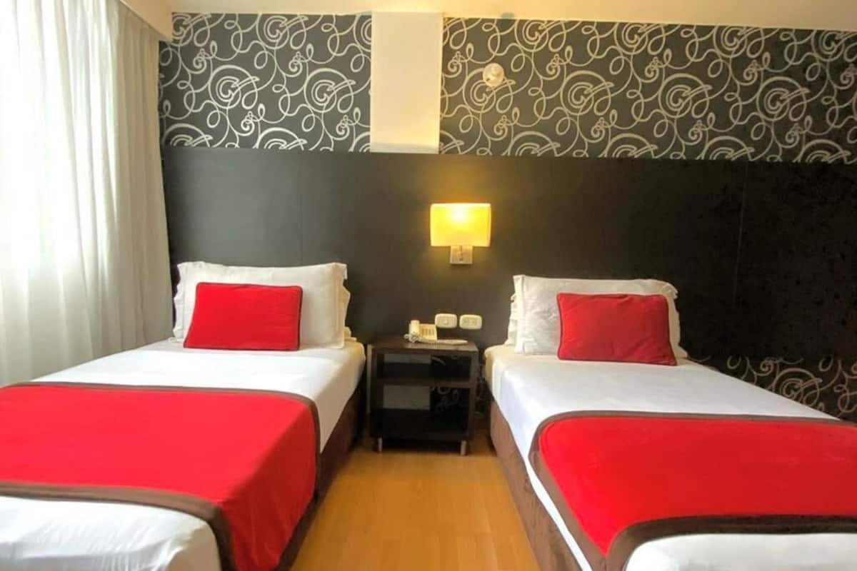 Quarto de hotel com duas camas, cada uma com uma almofada vermelha de destaque, uma mesa de cabeceira com abajur e papel de parede estampado.