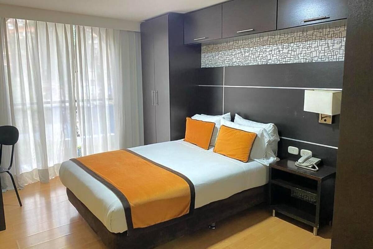 Quarto de hotel com uma cama grande, mesa de cabeceira com telefone e janela com cortinas.