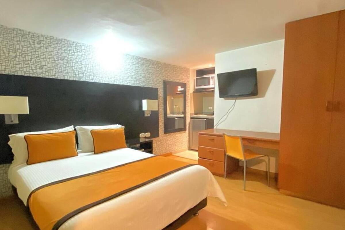 Quarto de hotel com cama, escrivaninha, cadeira, televisão e guarda-roupa.