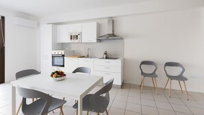Cozinha moderna com armários brancos, micro-ondas, forno e chaleira sobre a bancada. Há uma mesa de jantar com quatro cadeiras e uma fruteira em cima.