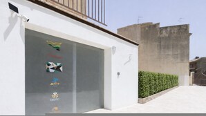 Um edifício moderno com fachada branca, porta de vidro com logotipos, parede de concreto e um pequeno jardim.