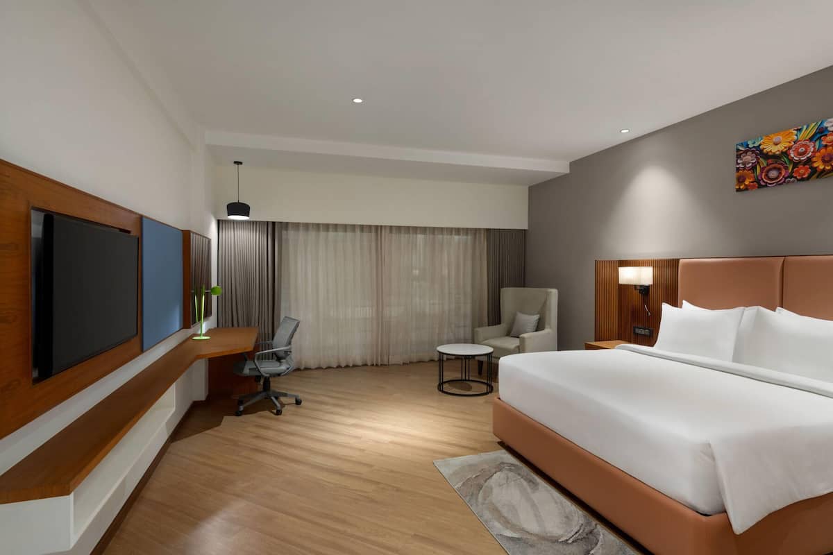 Quarto de hotel com uma cama grande, uma escrivaninha com cadeira, televisão, mesa pequena e janela com cortinas.