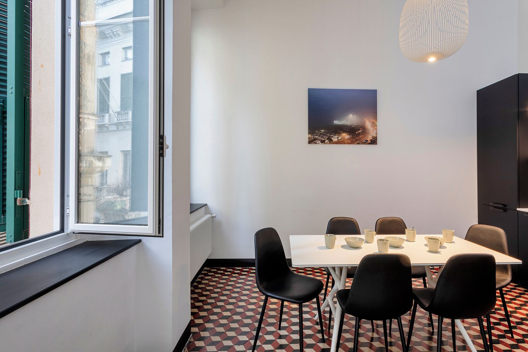 Uma área de refeições com mesa posta para quatro pessoas, cadeiras pretas, piso estampado, vista para a cidade pela janela e uma luminária pendente.