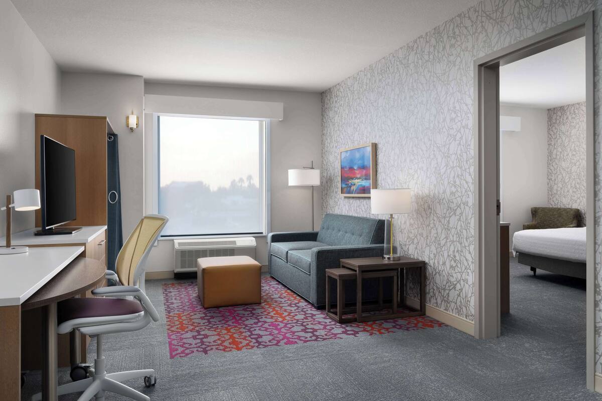 Quarto de hotel com escrivaninha, cadeira, sofá, cama e uma janela grande.