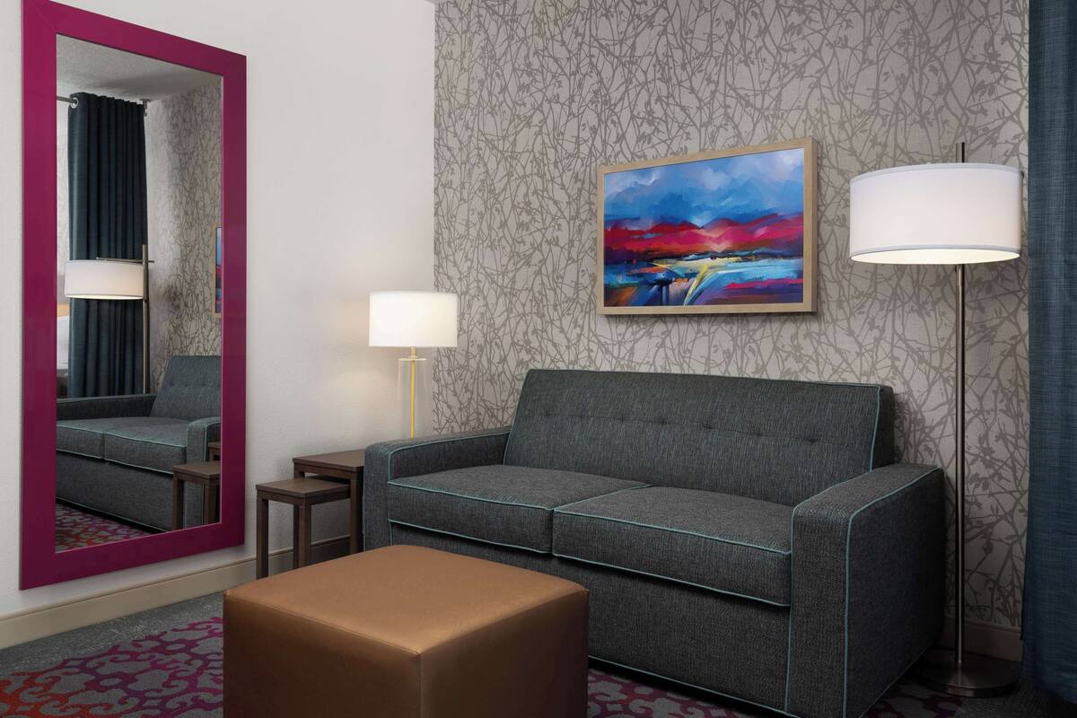 Quarto de hotel moderno com sofá cinzento, um espelho grande, um quadro abstrato emoldurado, um abajur e um tapete estampado.