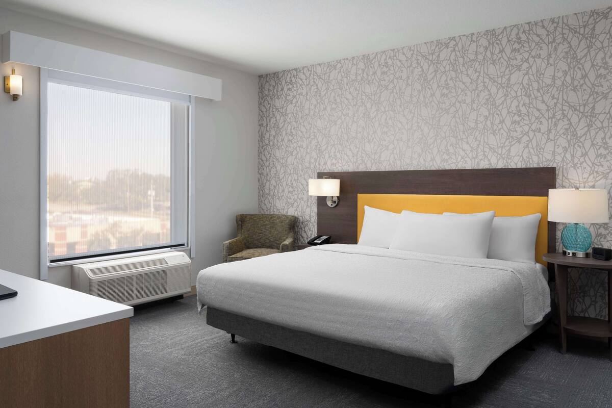 Quarto de hotel com uma cama grande, uma mesa, uma cadeira e uma janela com persianas.
