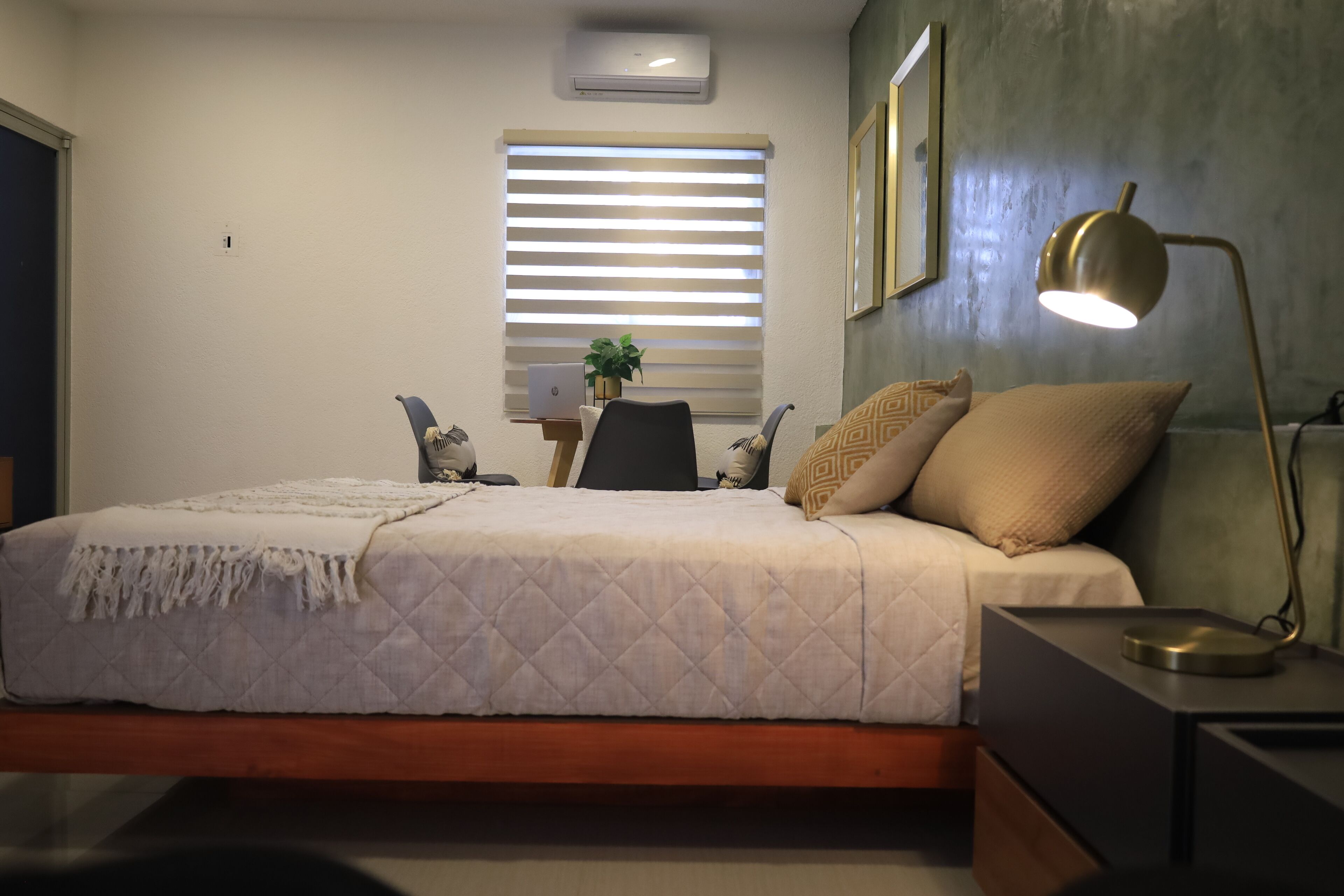 Quarto com cama, mesa de cabeceira, luminária, escrivaninha com cadeira e janela com persianas.