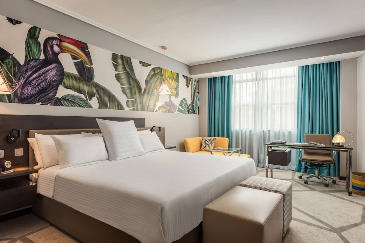 Quarto de hotel com uma cama grande, uma escrivaninha, uma cadeira e um mural na parede com um tucano.