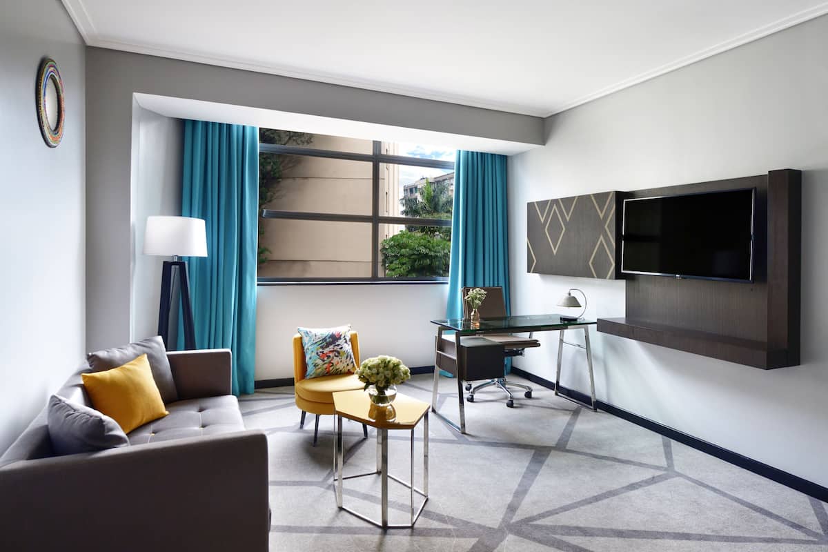 Quarto de hotel moderno com um sofá cinzento, uma pequena mesa com um vaso, uma escrivaninha, uma cadeira e uma televisão na parede.
