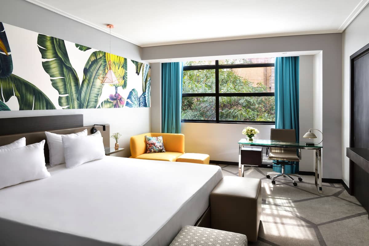 Quarto de hotel com cama grande, escrivaninha, cadeira e janela com cortinas.