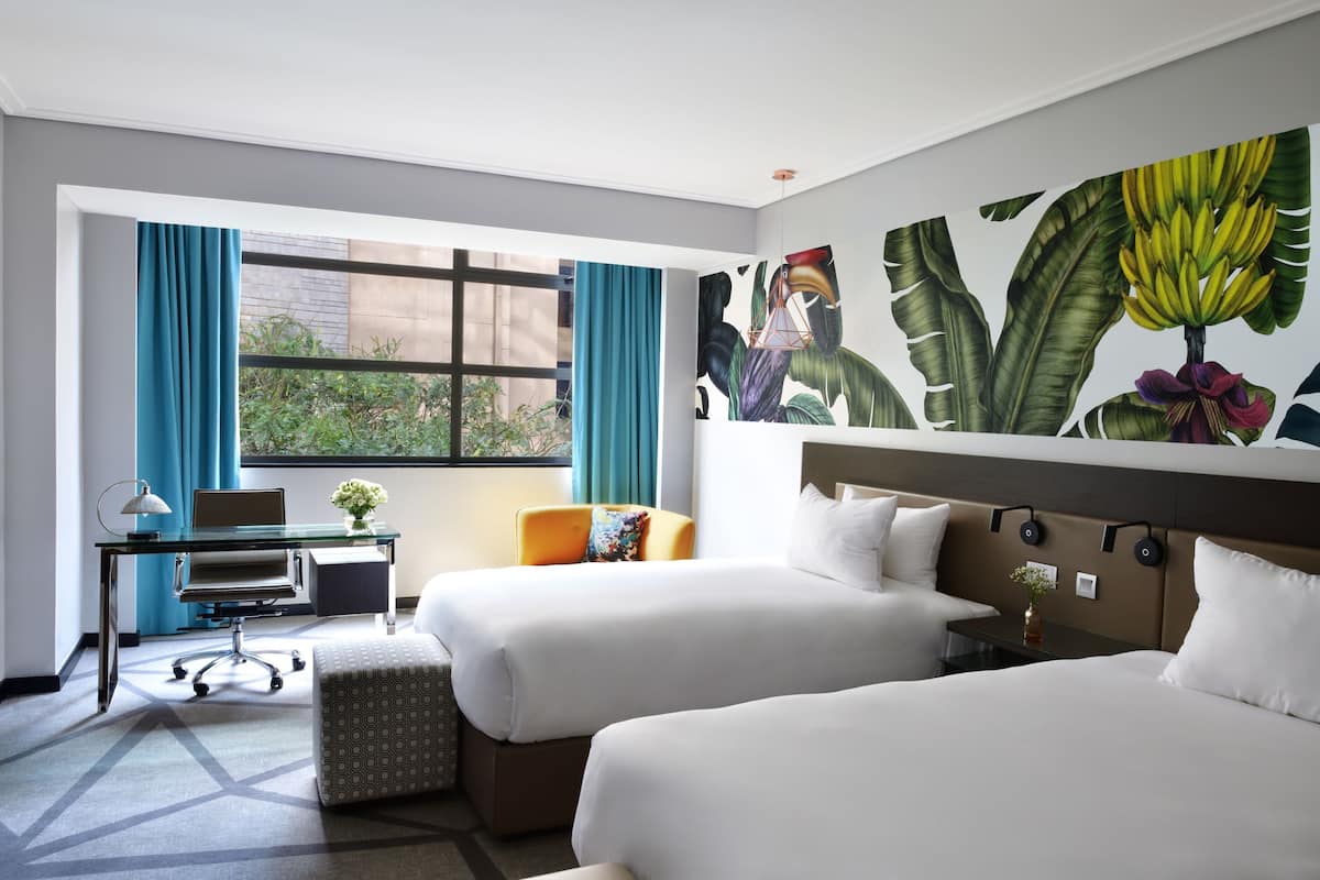 Quarto de hotel com duas camas, uma escrivaninha, uma cadeira e uma janela grande com cortinas.