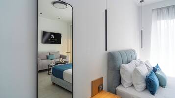 Quarto de hotel moderno com um espelho grande, uma cama com almofadas brancas e azuis, um sofá cinzento, uma mesa pequena e uma televisão de tela plana na parede.