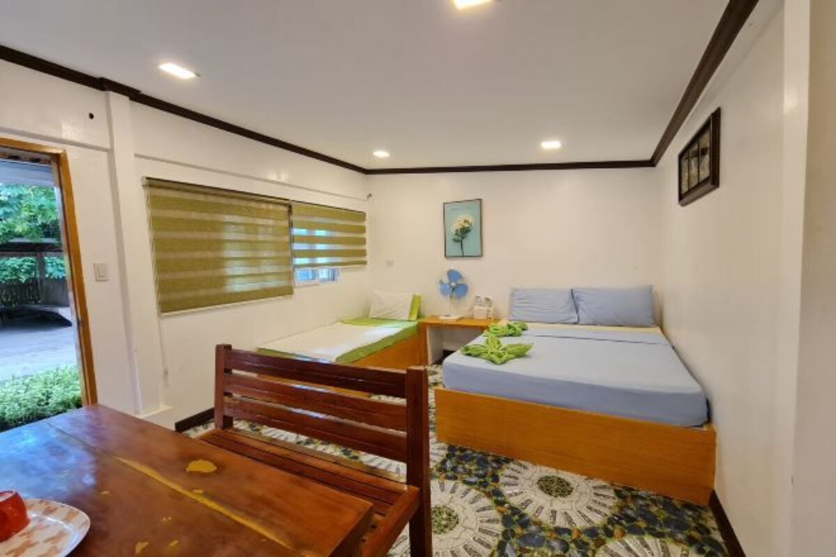Quarto pequeno e compacto com uma cama de solteiro, uma mesa de madeira com um prato e uma chávena, um banco e uma janela com persianas.
