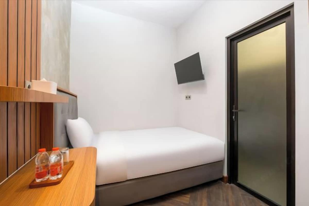 Quarto pequeno e moderno com uma cama, uma mesinha de madeira com garrafas, uma TV fixada na parede e uma porta deslizante.