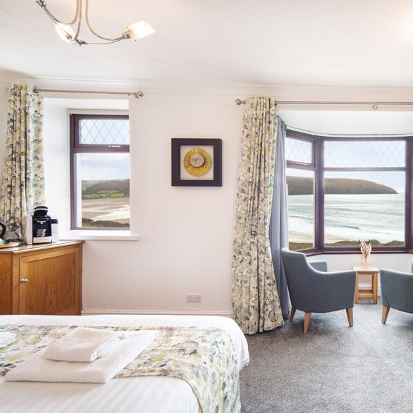 Quarto com cama, uma mesinha com luminária, uma cadeira e vista para o mar através de grandes janelas.
