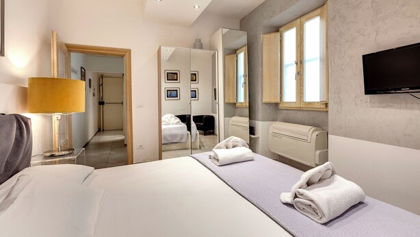 Quarto de hotel com cama, luminária, televisão, espelho e janela.