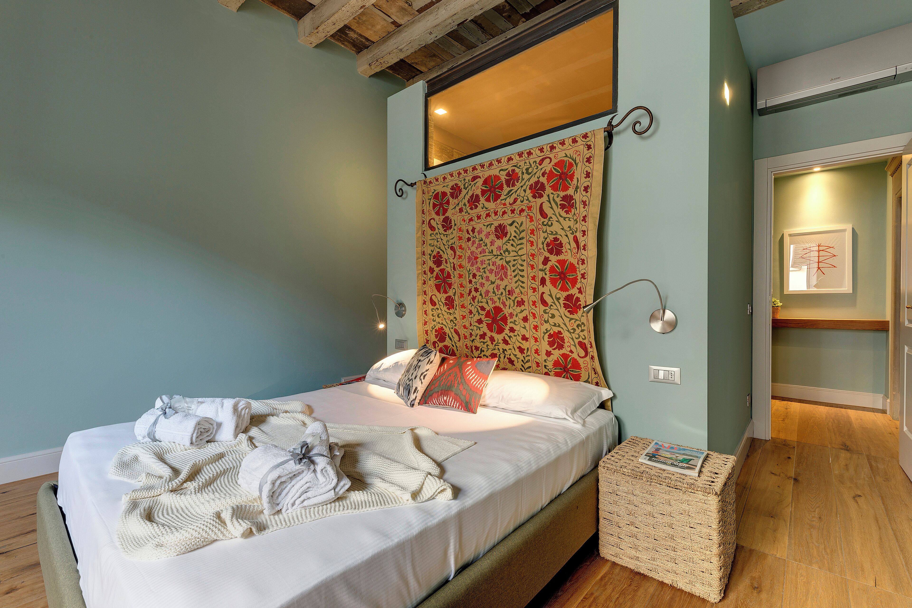 Cama bem arrumada com toalhas e um edredom, cabeceira decorativa, luminária de cabeceira e uma pequena cesta no chão.