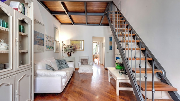 Uma sala de estar ampla com escada de madeira, móveis brancos e uma estante de livros.
