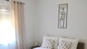 Um quarto com cama de madeira, roupa de cama branca, almofadas florais, uma mesa de cabeceira, um abajur e uma obra de arte emoldurada na parede.