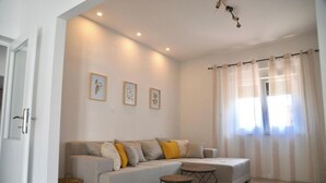 Uma sala de estar moderna com um sofá, duas mesinhas e uma janela grande com cortinas leves.
