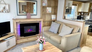 Uma sala de estar moderna com lareira, um sofá, uma mesa de centro com flores, uma TV, estantes de madeira e um espaço de cozinha.