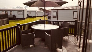 Um terraço com mesa e cadeiras, cercado por uma cerca de madeira, com vista para uma área gramada com casas móveis.