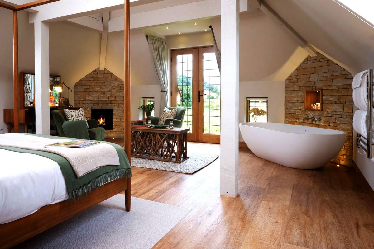 Um quarto com cama de dossel, lareira de pedra, uma janela grande e banheiro separado com banheira de imersão.
