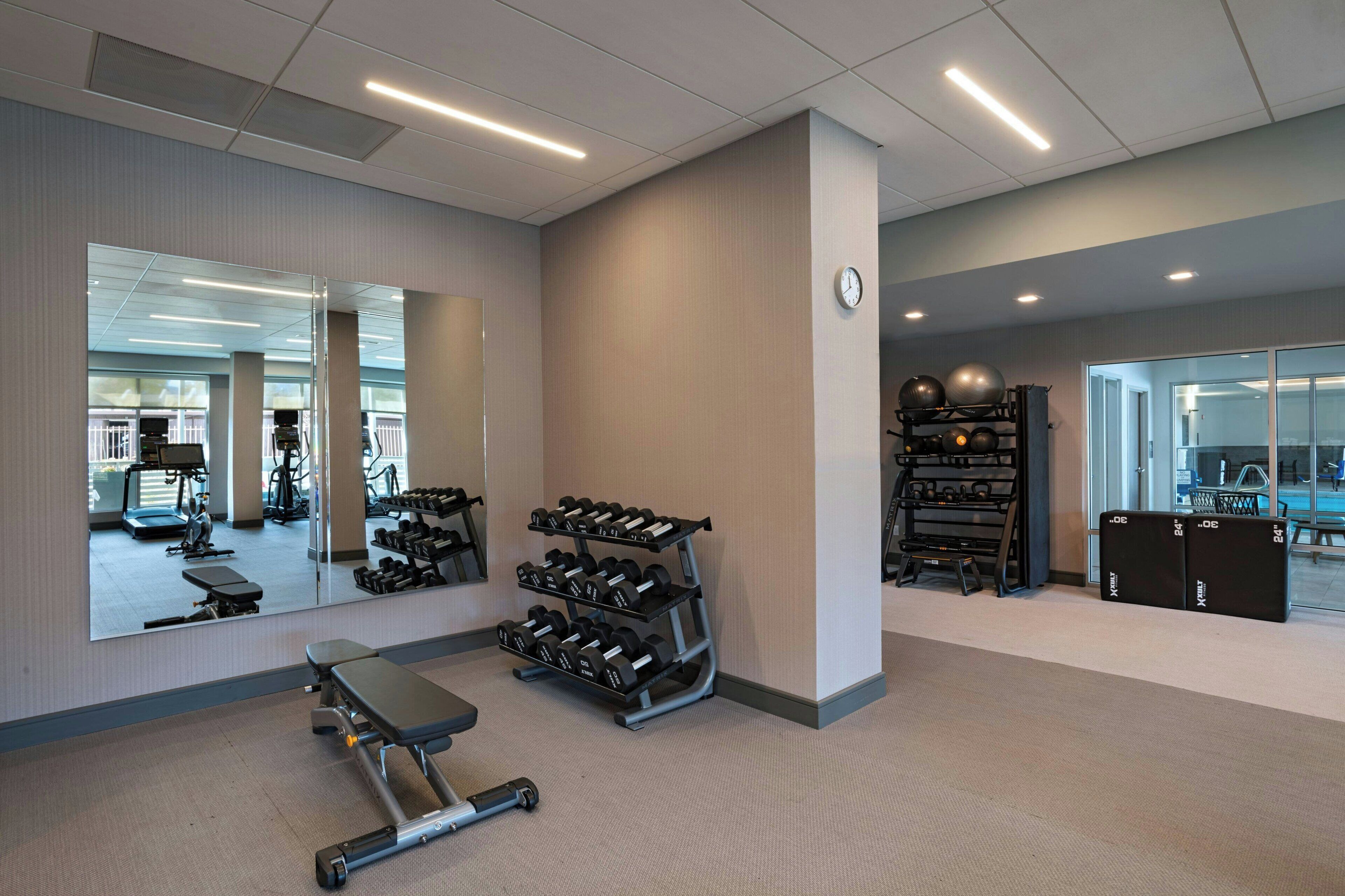 Uma academia com equipamentos de exercício, um banco e um espelho refletindo o ambiente.