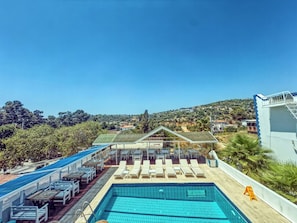 Piscina no terraço com espreguiçadeiras e mesas, cercada por vegetação e casas ao fundo.