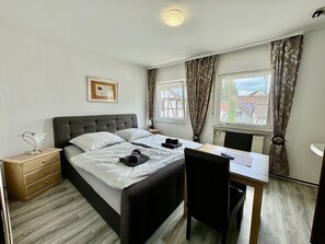 Quarto com cama, mesa de cabeceira, cadeira, escrivaninha e janelas com cortinas.