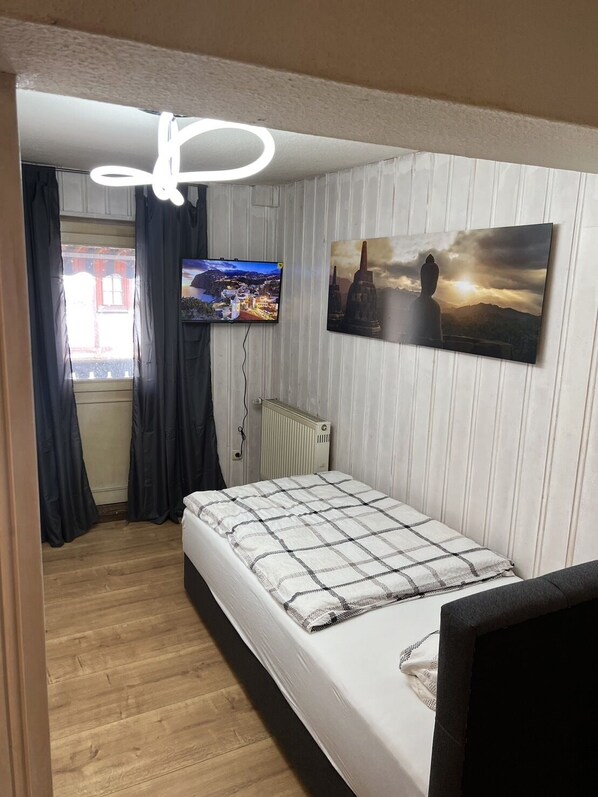 Quarto com cama, TV fixada na parede, janela com cortinas e radiador.