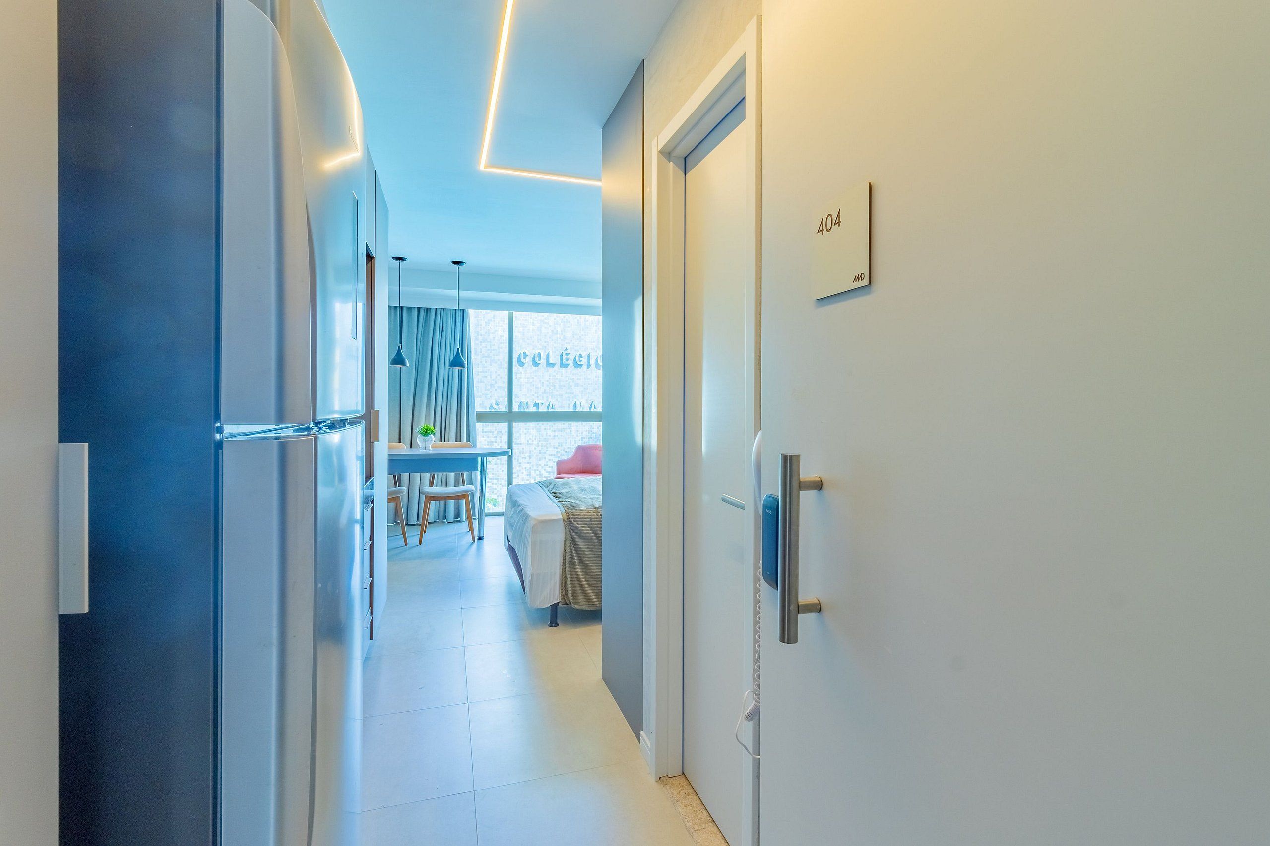 Corredor moderno de hotel com área de refeições, uma cama e porta com número de quarto.