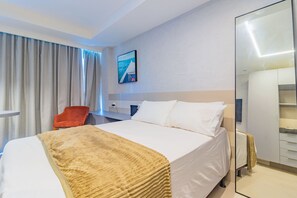 Quarto de hotel com cama, uma cadeira vermelha, escrivaninha, espelho e um quadro na parede.