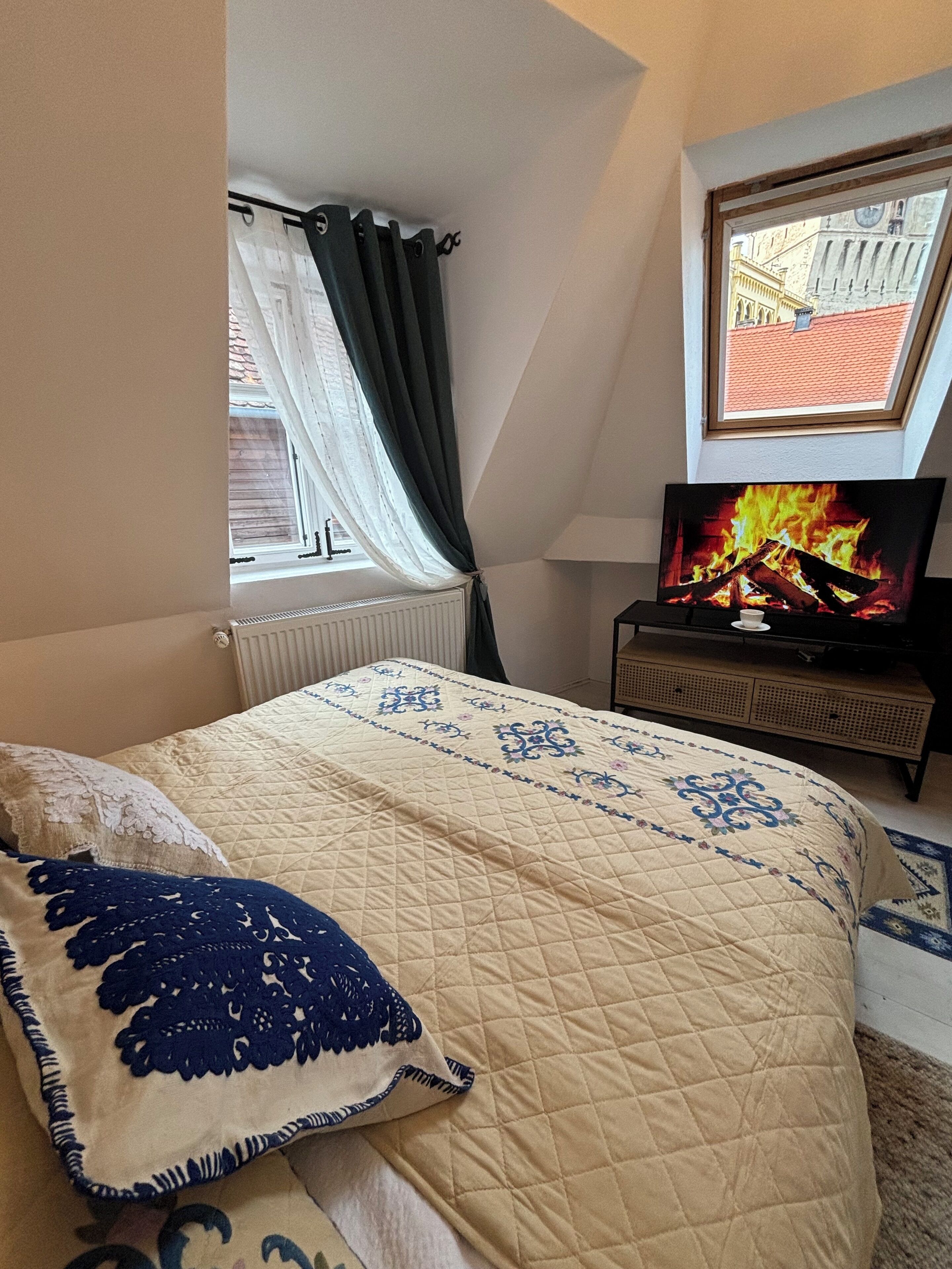 Quarto com cama, televisão exibindo uma lareira, janela com cortinas e vista para um edifício lá fora.