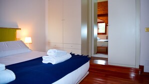 Cama bem arrumada com roupa de cama branca e manta azul, abajur e guarda-roupa com espelho.