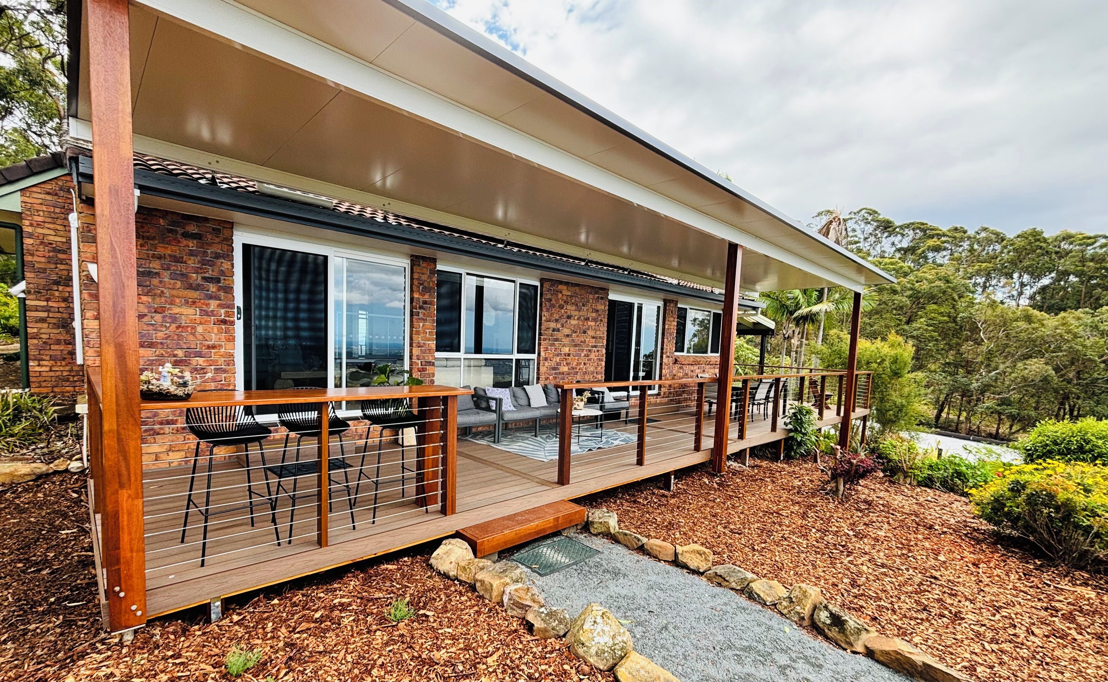 Uma casa moderna com deck de madeira, grandes janelas e vista para o verde ao redor.