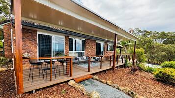 Uma casa moderna com deck de madeira, grandes janelas e vista para o verde ao redor.