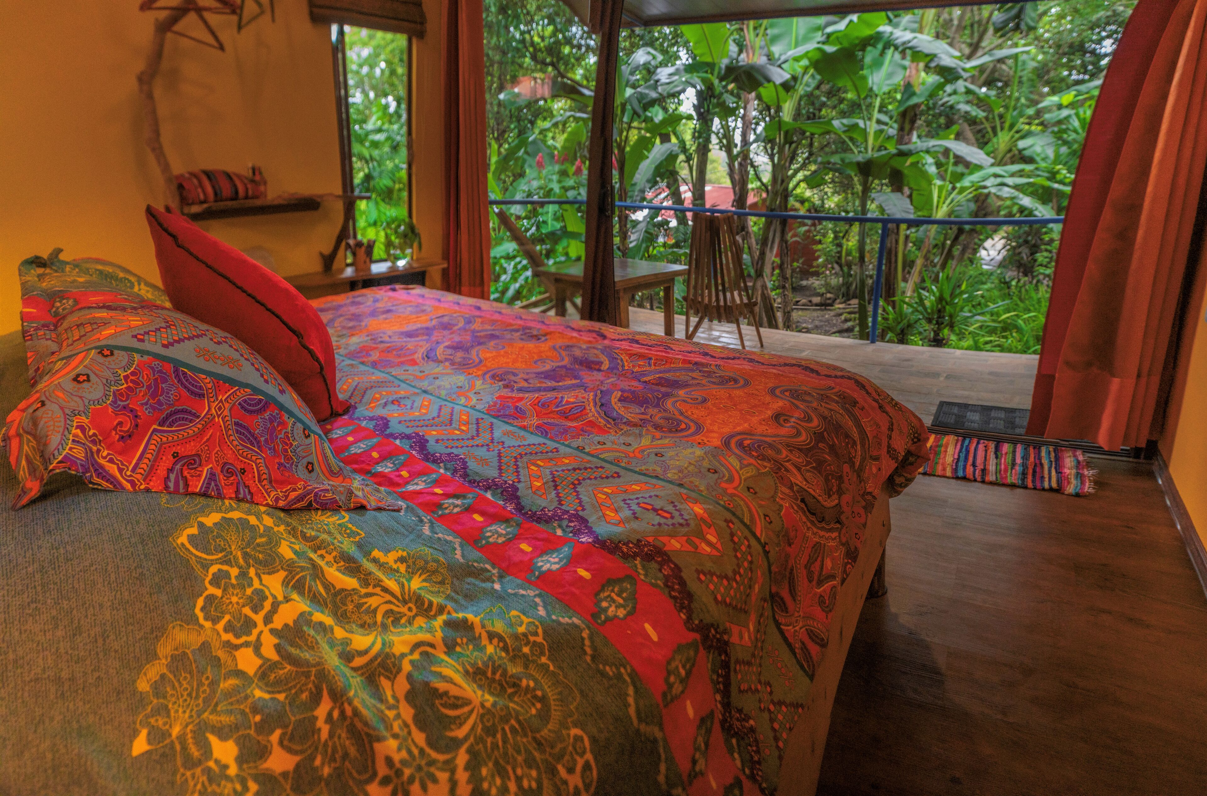 Uma cama com roupa de cama colorida, vista de vegetação exuberante através de uma varanda, e piso de madeira.