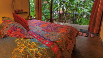 Uma cama com roupa de cama colorida, vista de vegetação exuberante através de uma varanda, e piso de madeira.