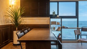 Uma sala de jantar com uma mesa comprida, cadeiras, um vaso de planta e vista para o mar através de grandes janelas.