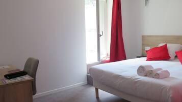 Quarto de hotel com uma cama, uma escrivaninha, uma cadeira, cortina vermelha e uma pequena planta na parede.