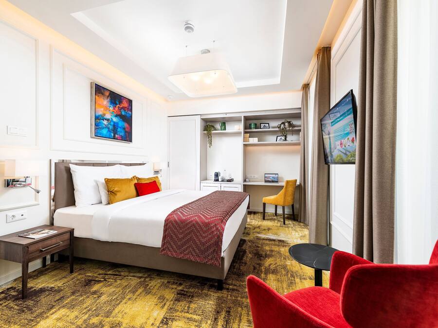 Quarto de hotel com cama, escrivaninha, cadeira e televisão.