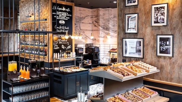 Um café com balcão de café, prateleiras com bebidas e uma variedade de pastéis e sanduíches expostos em mesas de madeira.