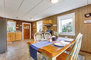 Uma área de refeições com uma mesa de madeira posta para quatro pessoas, rodeada por cadeiras brancas. Uma cozinha é visível ao fundo, com armários de madeira e uma janela.