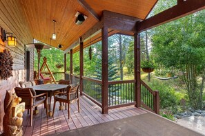 Um deck de madeira com mesa e cadeiras, cercado por corrimão, com vista para uma área arborizada.