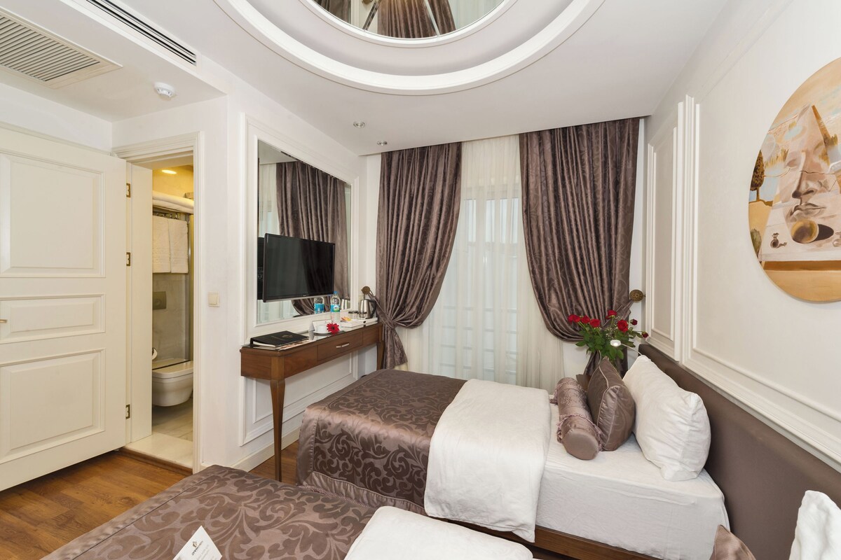 Quarto de hotel com uma cama, uma escrivaninha com computador, um espelho e uma janela com cortinas.