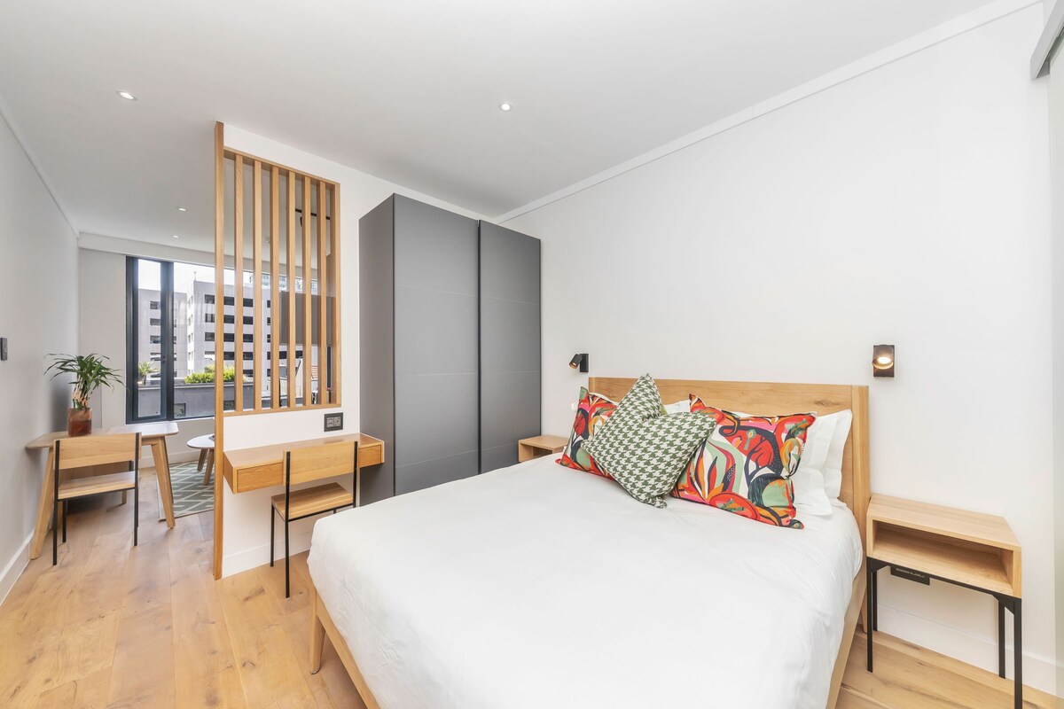 Quarto moderno com uma cama grande, uma escrivaninha e uma cadeira. Há uma janela com vista para edifícios.