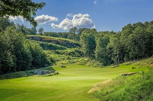 Um campo de golfe com haste de sinalização, rodeado por floresta densa e colinas onduladas.